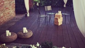 Um pátio externo com piso de madeira, parede de tijolos e uma pérgola com luzes de cordão. Há duas cadeiras e uma mesa com velas.