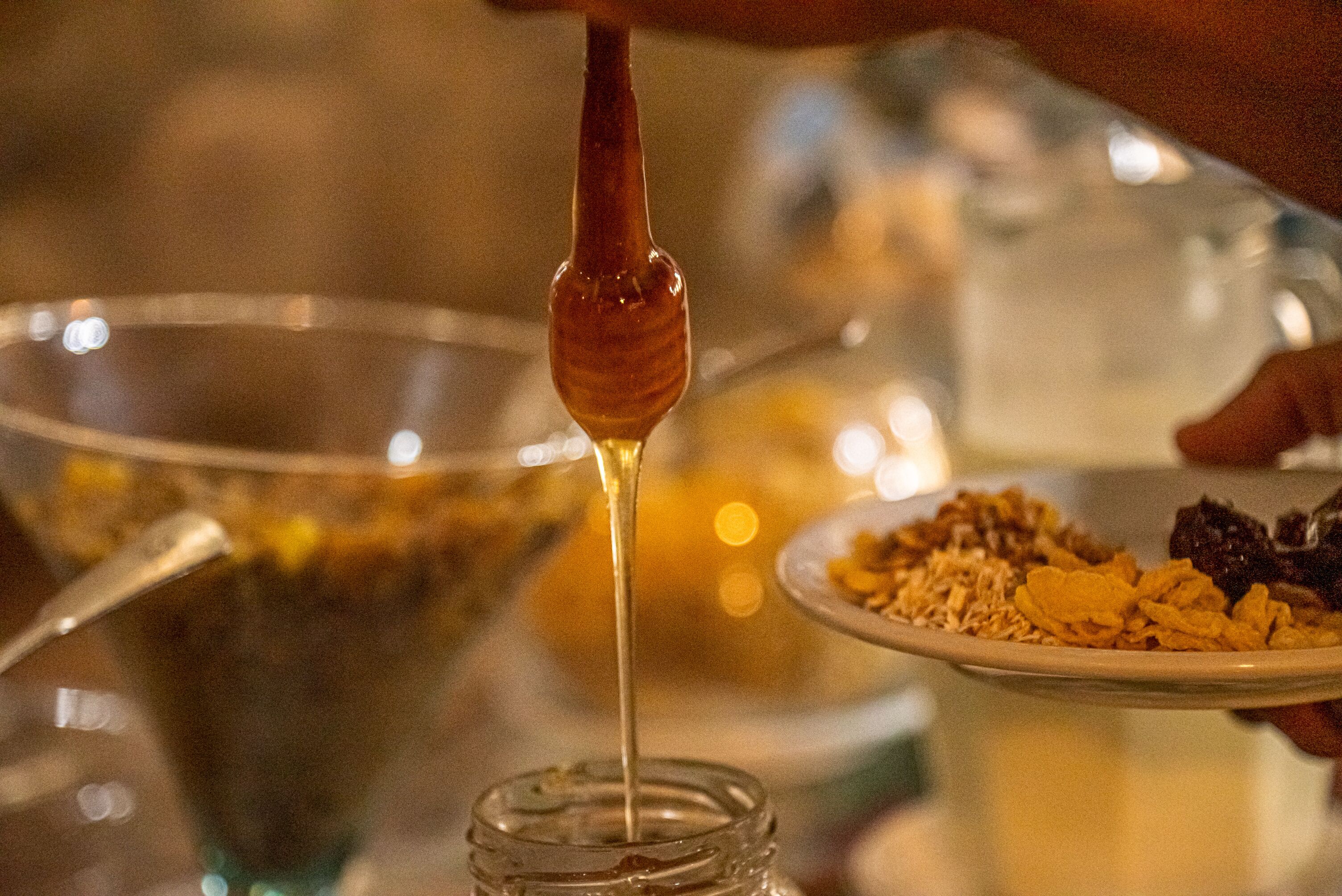 Um close-up de um colher de mel pingando mel em um pote, com uma tigela de granola e frutas secas ao fundo.