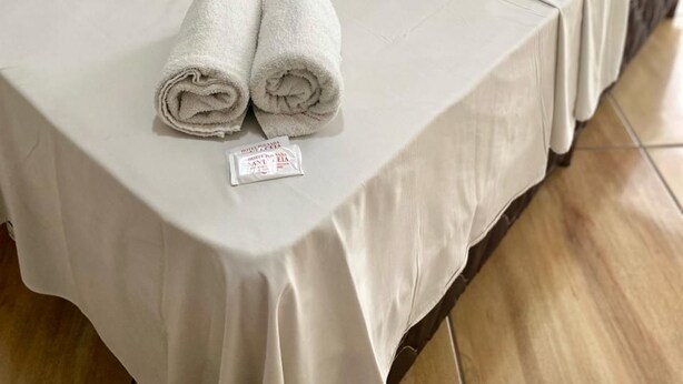 Cama bem arrumada com colcha branca e duas toalhas enroladas ao lado.