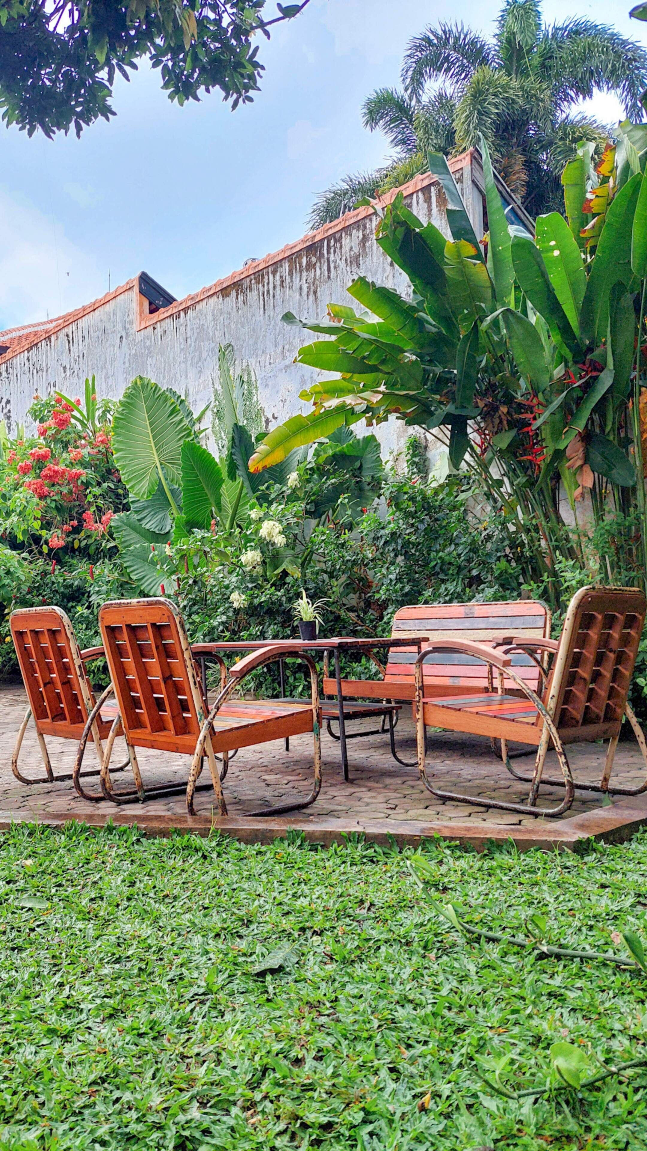 Um pátio com mesa e cadeiras de madeira, rodeado por vegetação exuberante e uma parede branca.