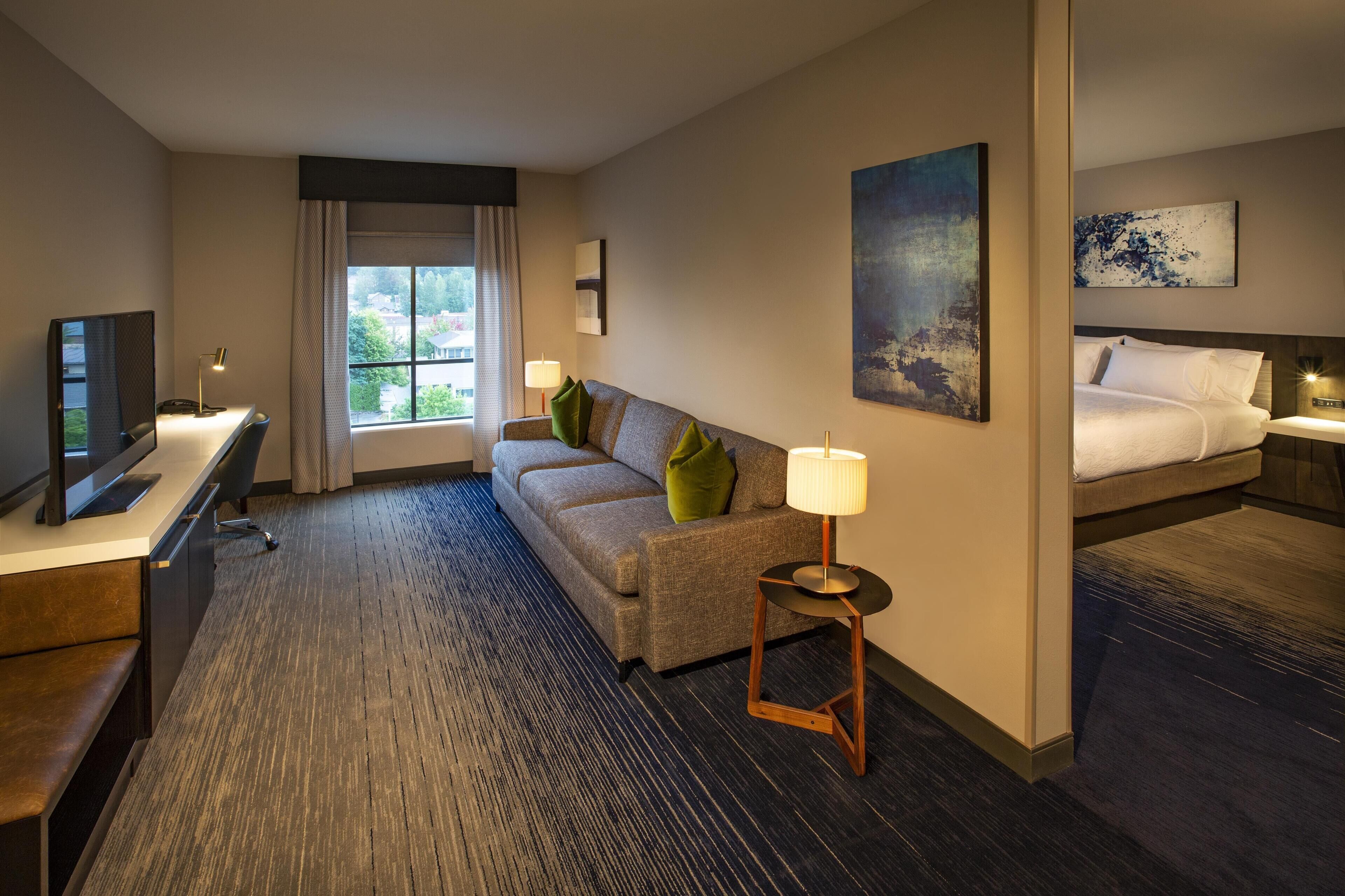 Quarto de hotel com escrivaninha, sofá, cama, janela com cortinas e obras de arte nas paredes.