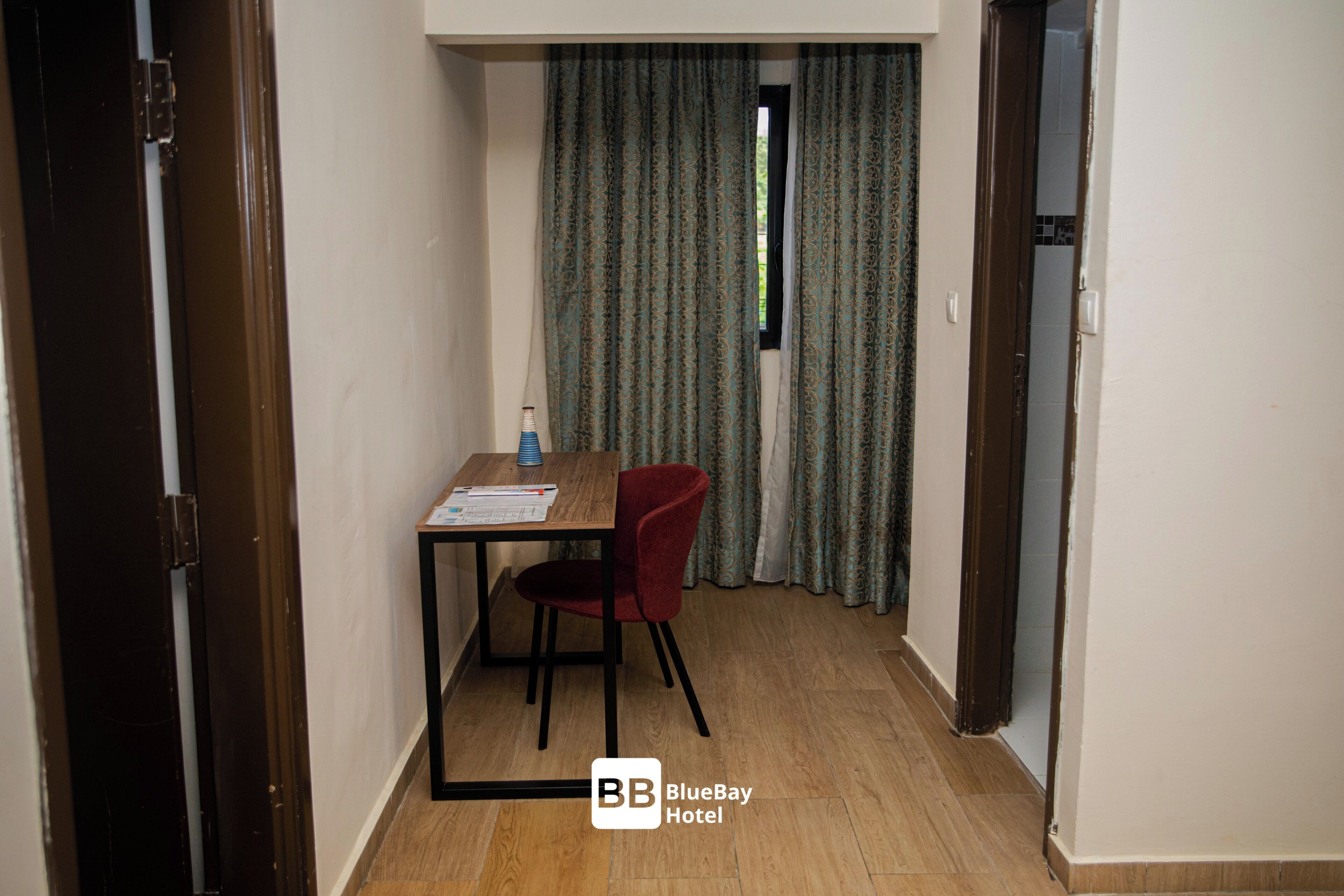 Um quarto pequeno com uma mesa de madeira, uma cadeira vermelha e uma janela com cortinas estampadas.
