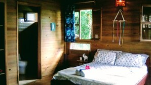 Quarto de cabana de madeira com cama, janela com cortinas, uma pequena mesa com uma xícara e um lustre.