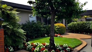 Um jardim bem cuidado com diversas plantas e flores, uma entrada com caminho sinuoso e um edifício com telhado de cerâmica.
