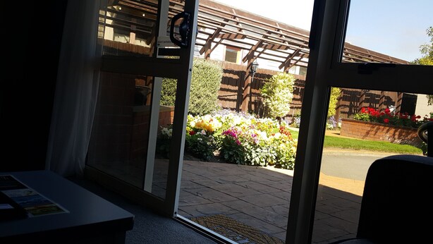 Quarto com porta de correr de vidro que dá para um alpendre externo com pérgola de madeira e diversas flores.