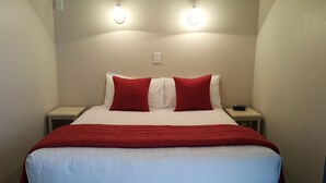Cama bem arrumada com manta vermelha e almofadas, duas mesas de cabeceira com abajures e uma parede lisa.