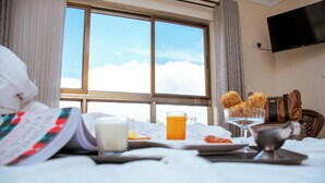 Quarto de hotel com cama, janela com vista para as nuvens, televisão fixada na parede e bandeja de café da manhã sobre a cama.