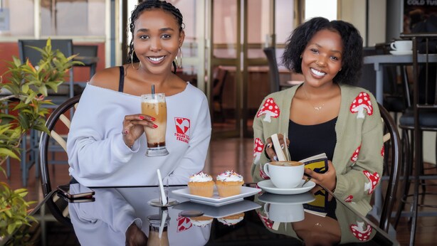 Duas pessoas sentadas à mesa com uma bebida, um cupcake e uma chávena, num café com cadeiras e uma planta ao fundo.