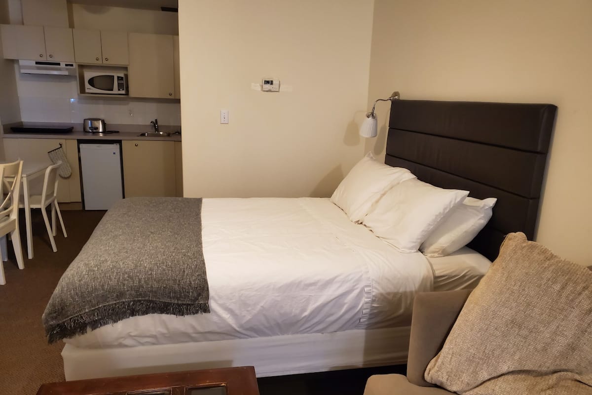 Quarto de hotel com cama, mesa de cabeceira, cadeira, cozinha compacta e uma pequena mesa.