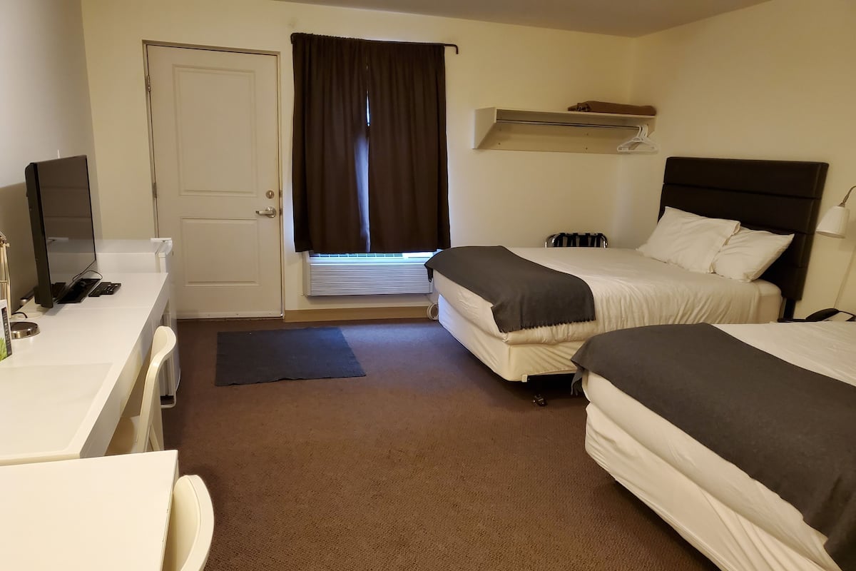 Quarto de hotel com duas camas, uma escrivaninha, uma cadeira, um computador e uma janela com cortinas.