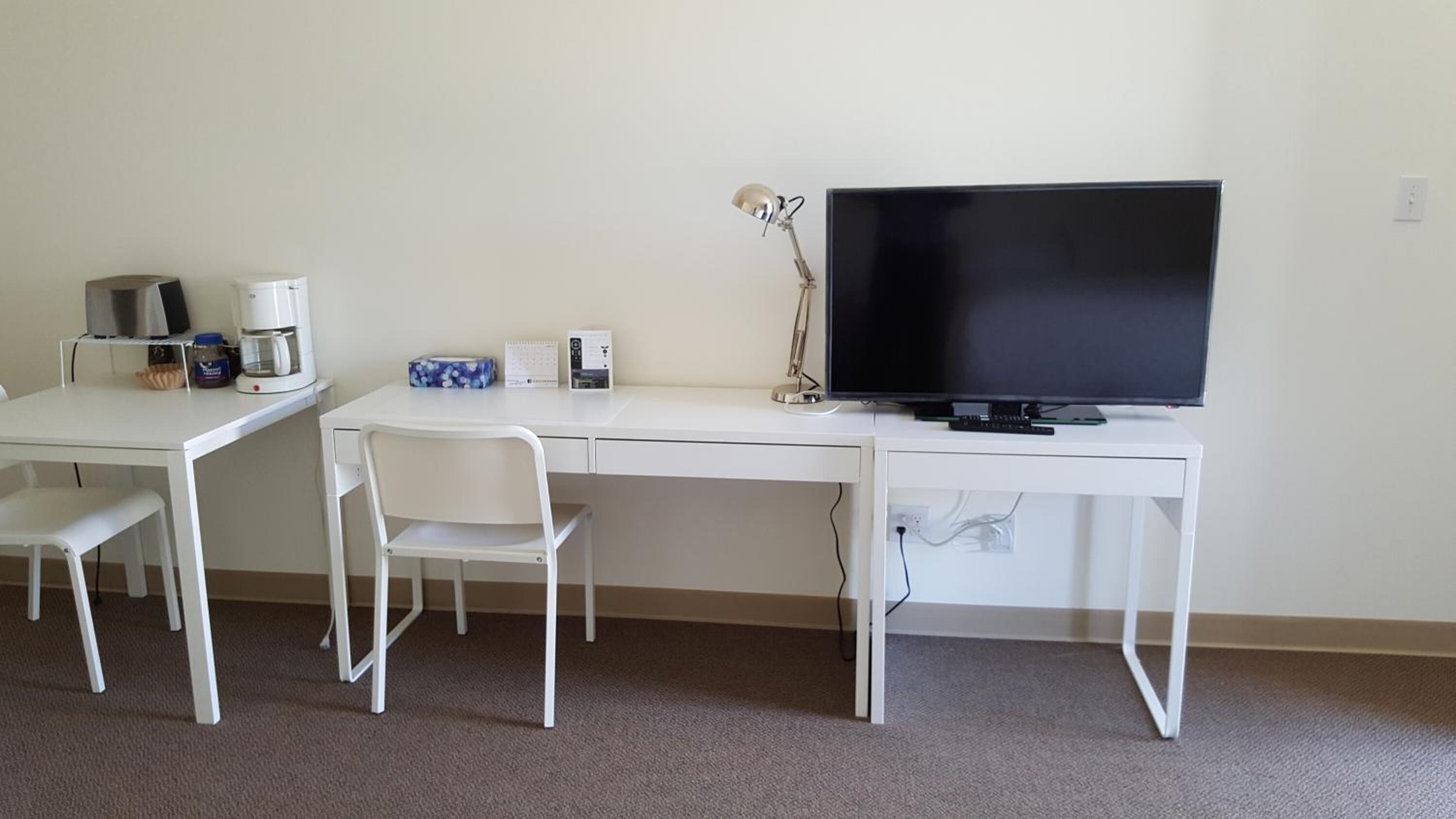 Quarto pequeno e limpo, com uma escrivaninha branca, uma cadeira, uma televisão e uma máquina de café.