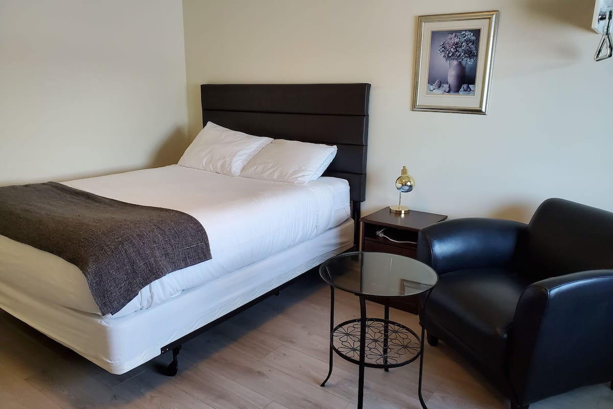 Quarto de hotel bem arrumado, com uma cama, uma mesa redonda pequena, uma cadeira e uma mesa de cabeceira com uma luminária.