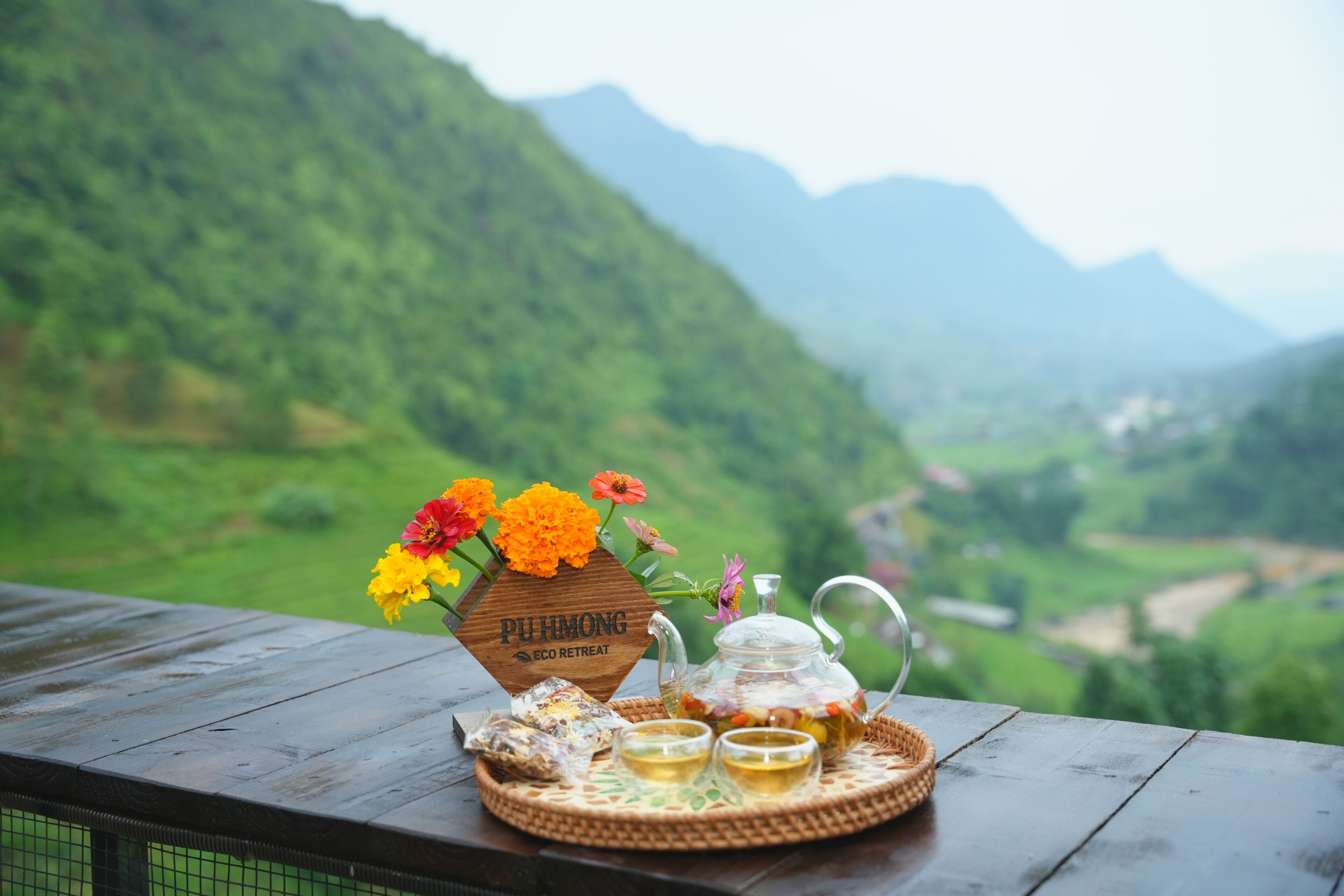 Uma mesa de madeira com uma bandeja de madeira que contém um frasco de vidro, uma chaleira e um vaso com flores.