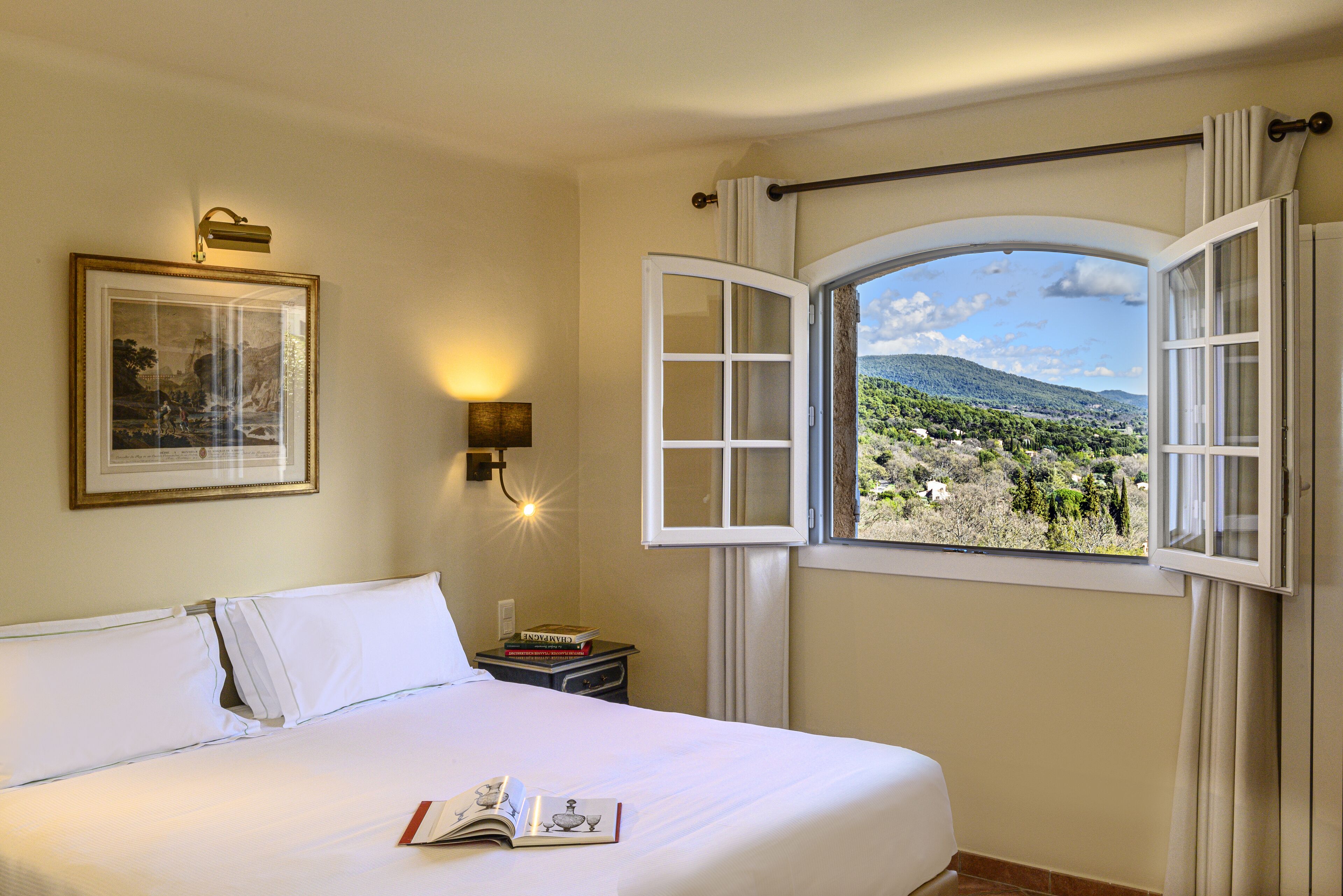 Quarto de hotel com uma cama, um quadro na parede, uma mesa de cabeceira com livros e uma janela com vista para uma paisagem montanhosa.