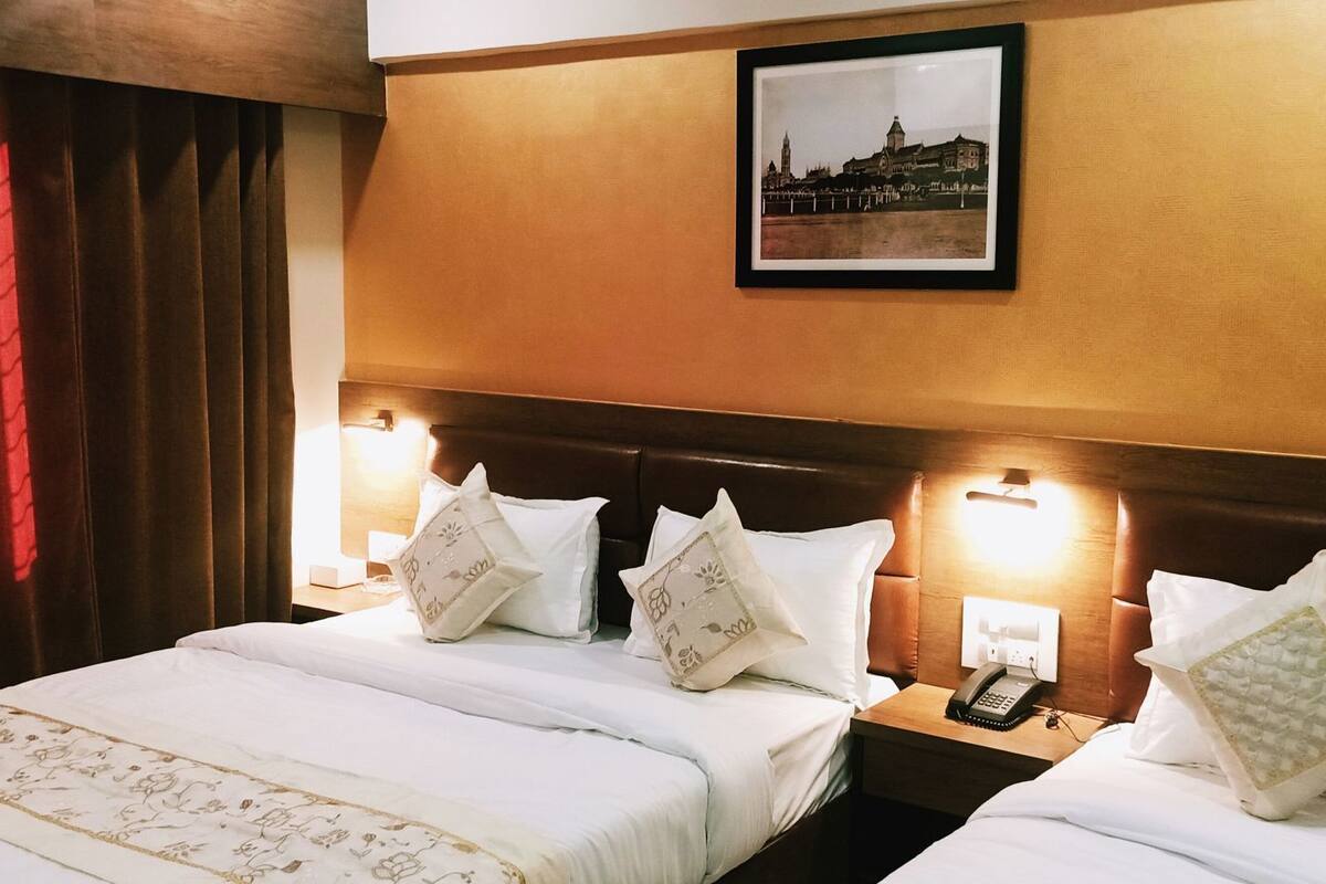 Quarto de hotel com duas camas, cabeceira de madeira, abajures, um quadro emoldurado na parede e uma cortina vermelha.
