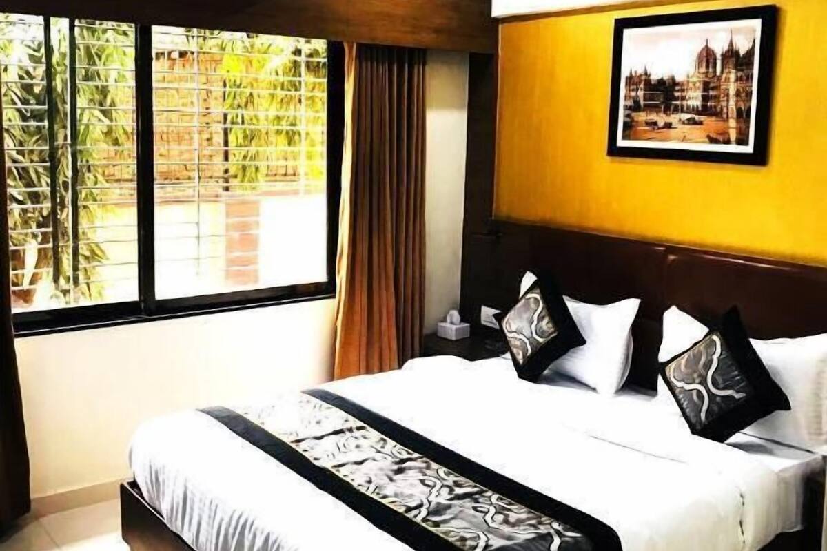 Quarto de hotel com cama, ar condicionado, ventoinha no teto, janela com cortinas e um quadro na parede.