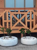 Uma estrutura de madeira com design geométrico, possivelmente um deck ou varanda, com dois vasos de pneus contendo plantas e pedras.