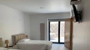 Quarto com duas camas, uma TV fixada na parede e vista para uma parede de pedra por uma porta aberta.
