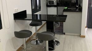 Cozinha moderna com micro-ondas, pia e pequena geladeira. Três banquetas com assentos pretos e bases cromadas. A cozinha possui bancada preta e painel de fundo trabalhado.