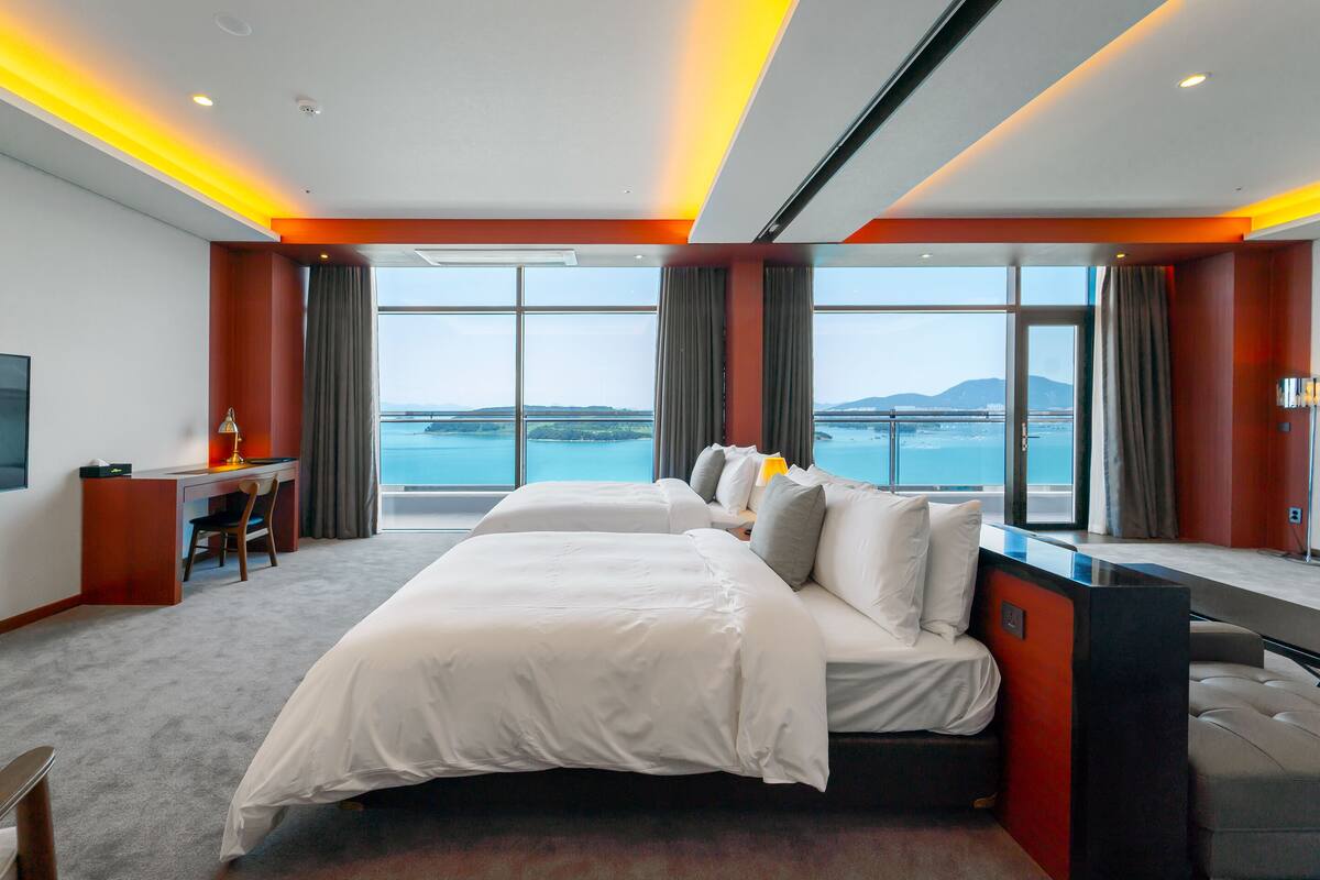 Quarto de hotel com uma cama grande, uma escrivaninha e uma TV. Tem vista para o mar e para as montanhas.