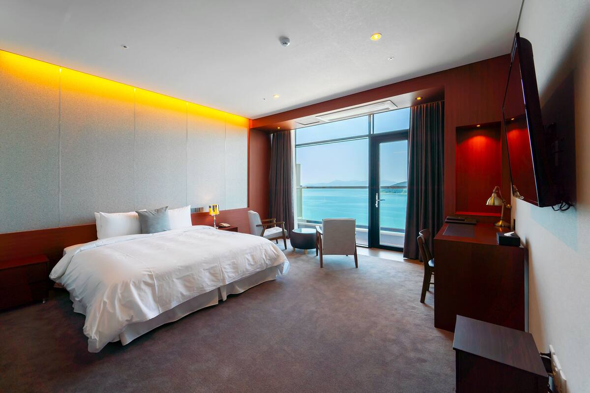 Quarto de hotel com cama grande, escrivaninha, cadeira e vista para o mar.