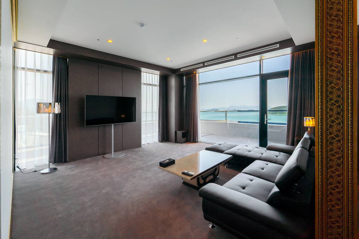 Uma sala de estar moderna com uma grande TV de tela plana, um sofá, uma mesa de centro e vista para o mar através de amplas janelas.