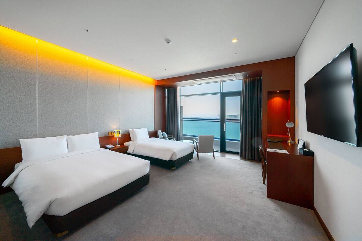 Quarto de hotel com duas camas, uma mesa, uma cadeira, uma TV e uma grande janela com vista para o mar.