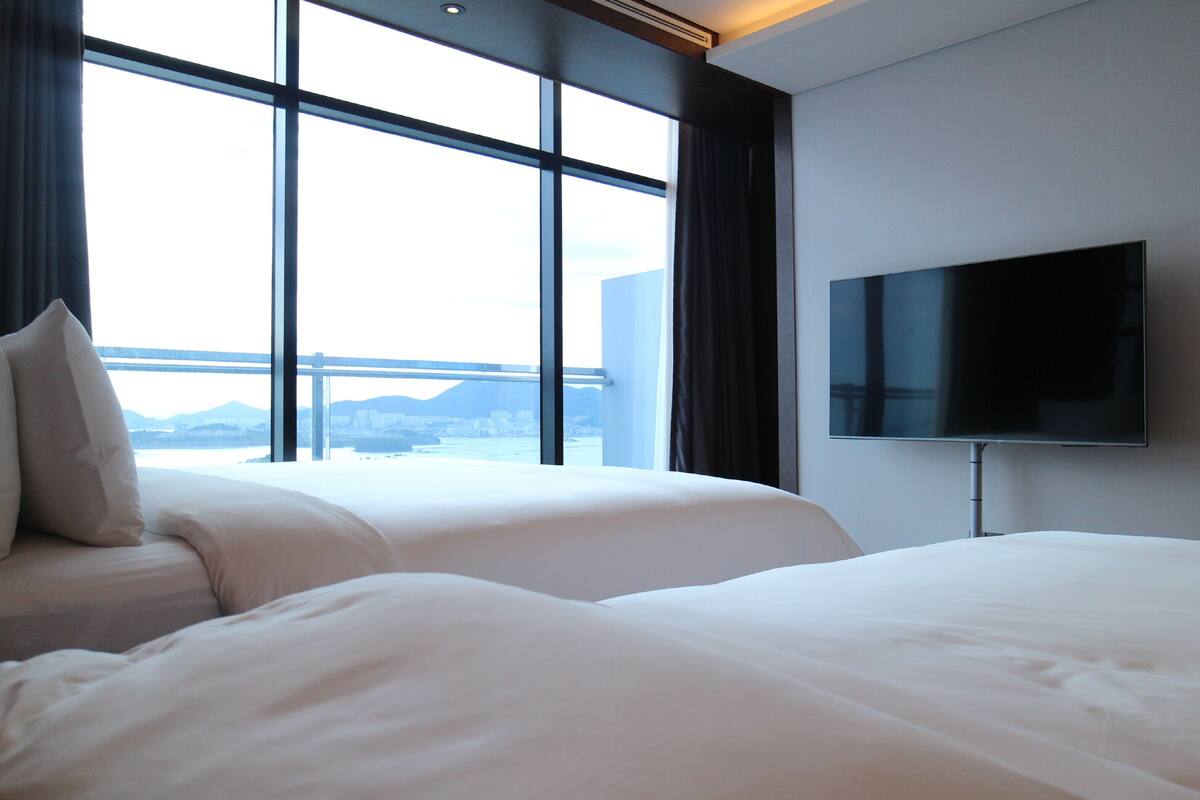 Quarto de hotel com duas camas, uma janela grande com vista para a cidade e uma televisão de tela plana fixada na parede.