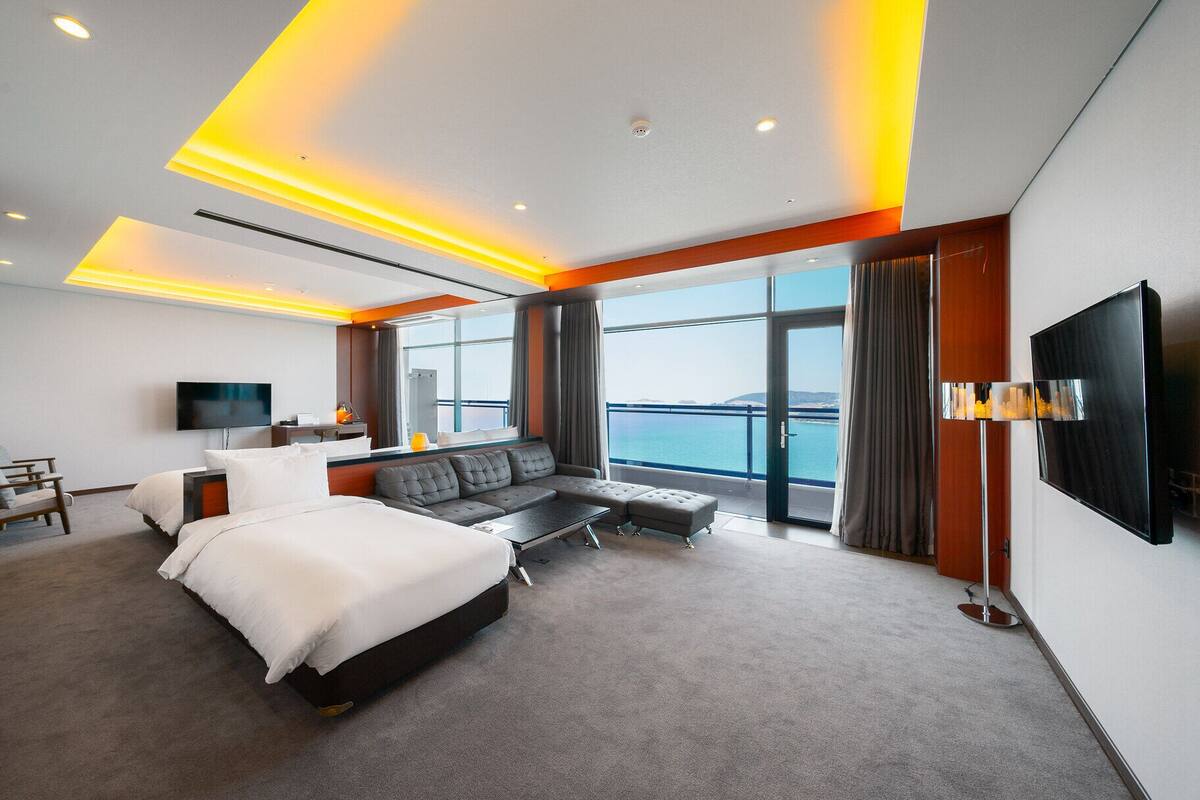 Quarto de hotel moderno com uma cama grande, um sofá, uma escrivaninha e uma televisão. O quarto possui uma varanda com vista para o mar.