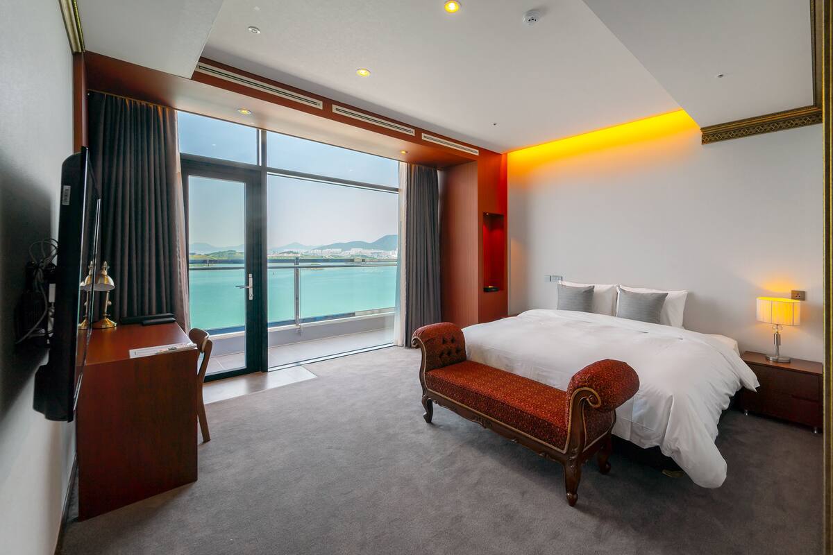 Quarto de hotel com cama grande, escrivaninha, cadeira, televisão e vista para o mar.