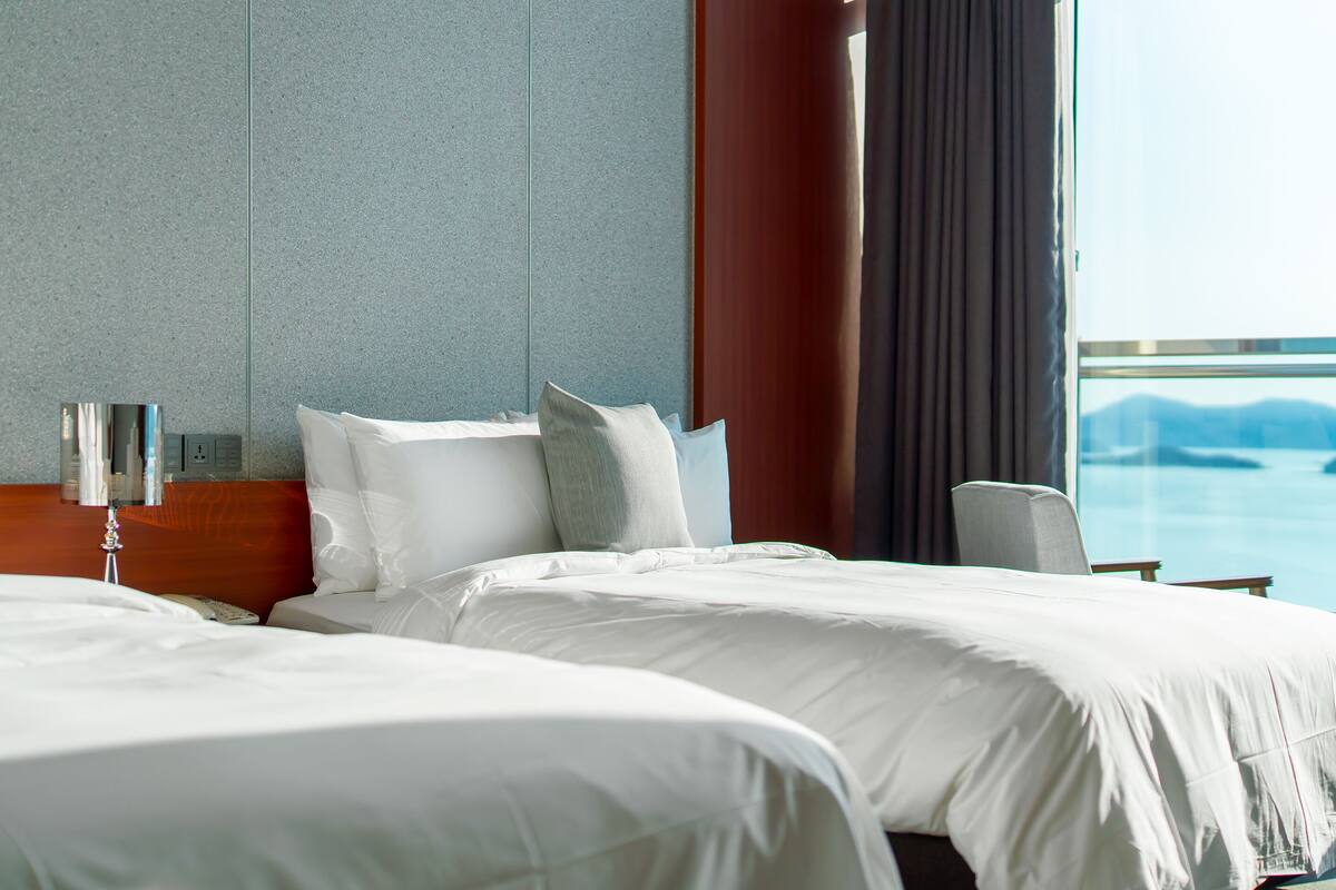 Quarto de hotel com duas camas, cabeceira de madeira, mesa de cabeceira e vista para o mar.