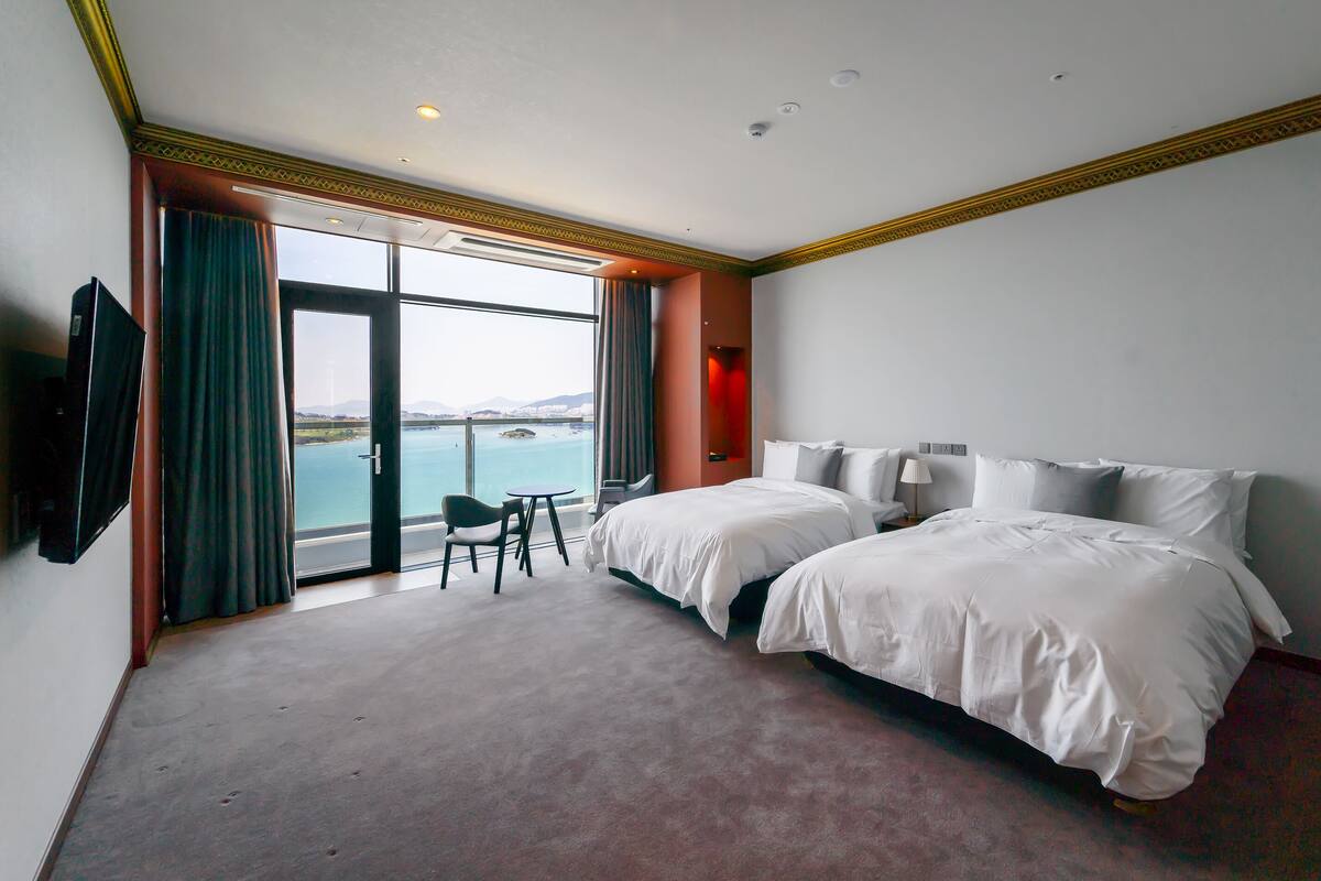 Quarto de hotel com duas camas, televisão, escrivaninha e vista para o mar.