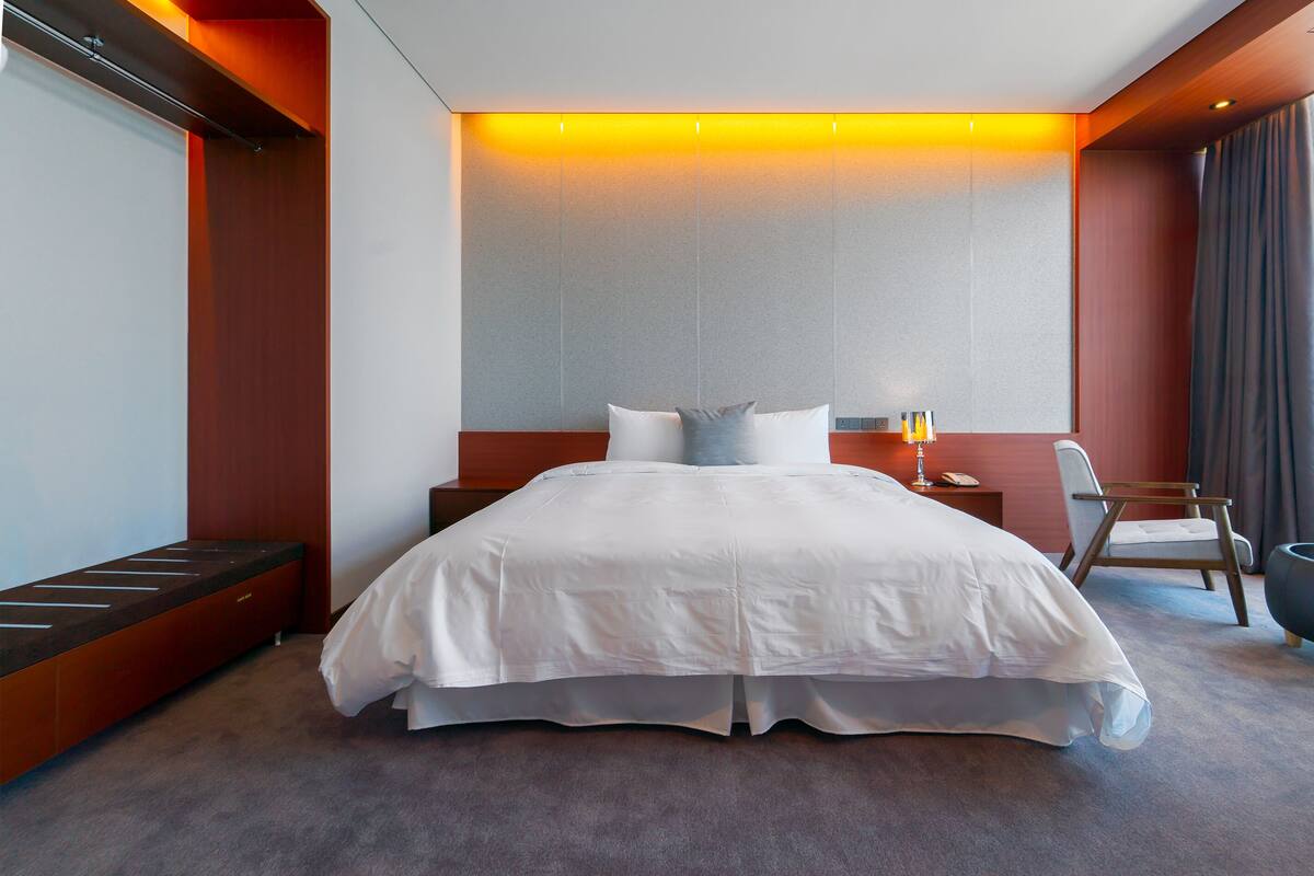 Quarto de hotel moderno com uma cama grande, mesa de cabeceira, uma cadeira e vista para a cidade.