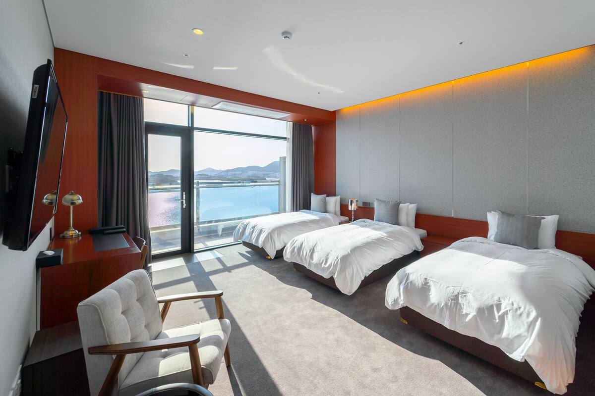 Quarto de hotel com duas camas, uma escrivaninha, uma cadeira, televisão e uma janela grande com vista para o mar.