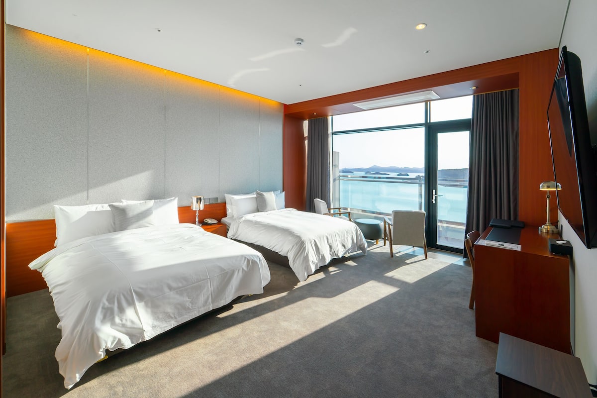 Quarto de hotel com duas camas, uma escrivaninha, uma cadeira, televisão e uma janela grande com vista para o mar.