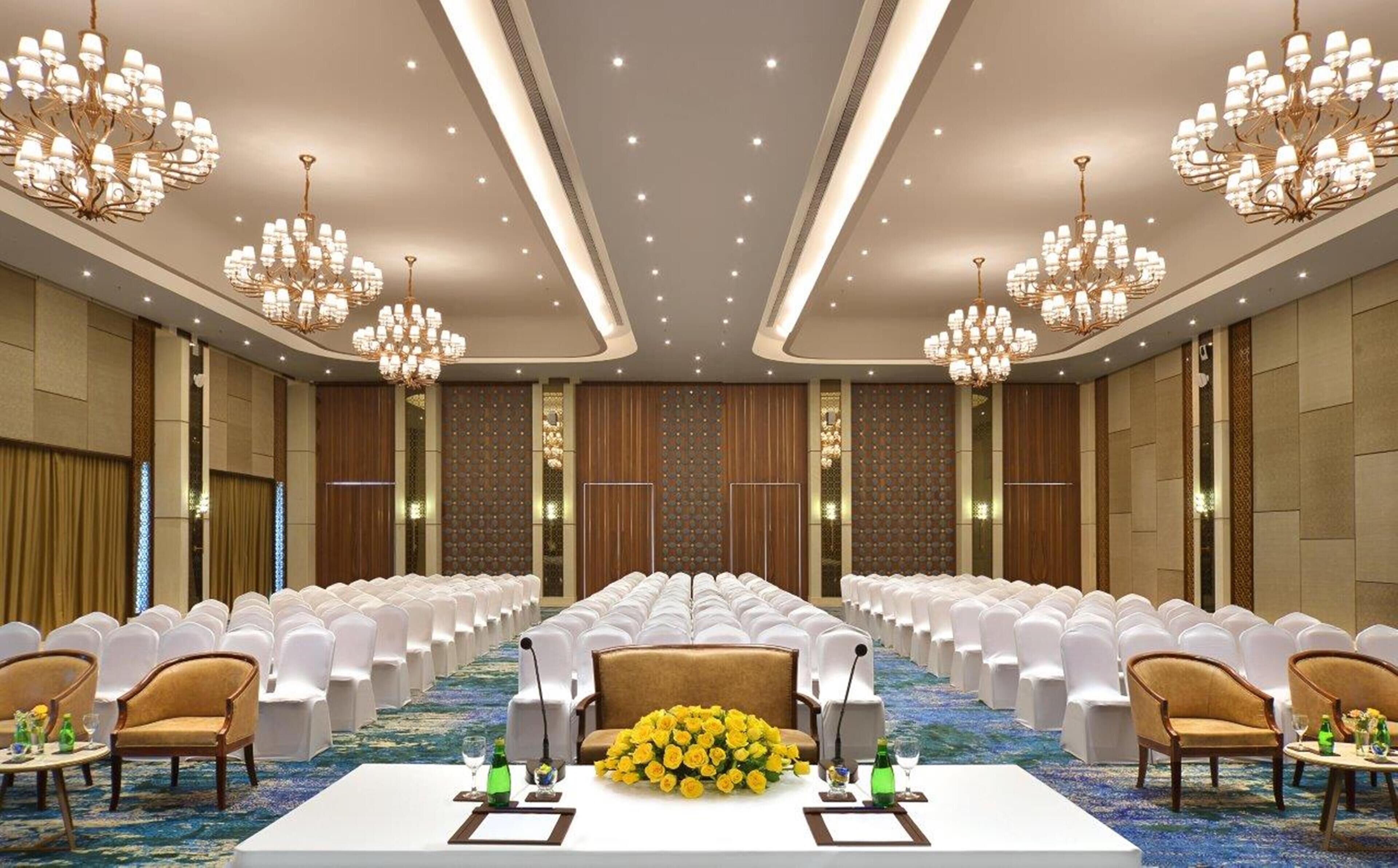 Sala de conferência com fileiras de cadeiras brancas, uma mesa comprida com microfones e lustres.