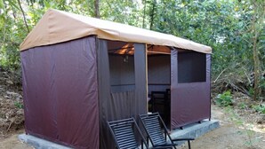 Uma pequena tenda de um único compartimento, com teto bege, porta e janela. Duas cadeiras de metal preto estão em frente à tenda.