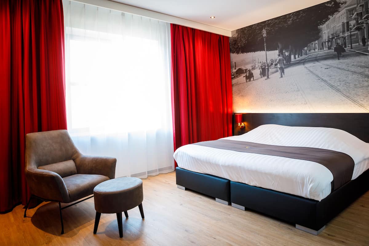 Quarto de hotel com uma cama grande, uma cadeira, um pequeno banquinho e uma luminária na parede.