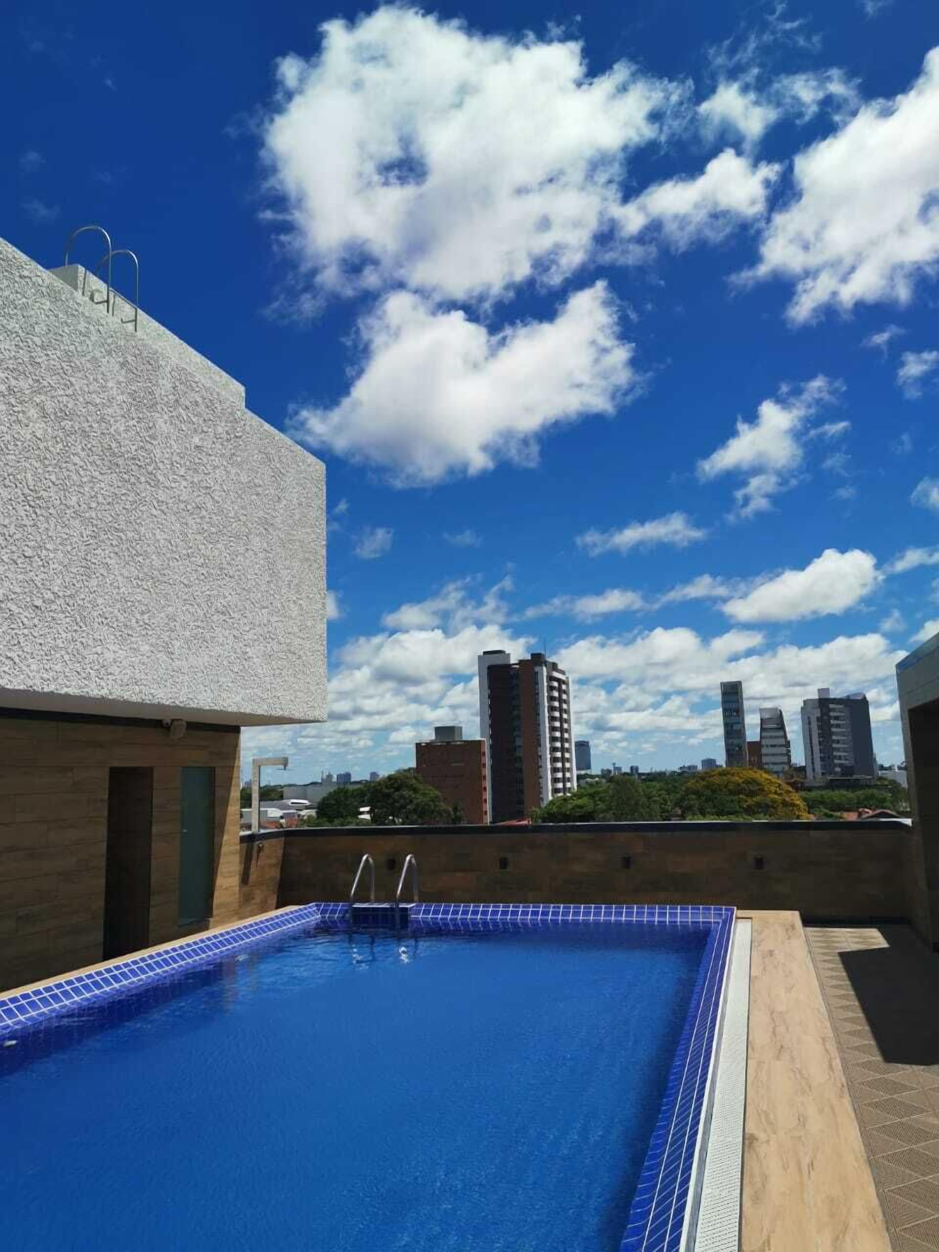 Piscina ao ar livre com céu azul e nuvens, cercada por edifícios.