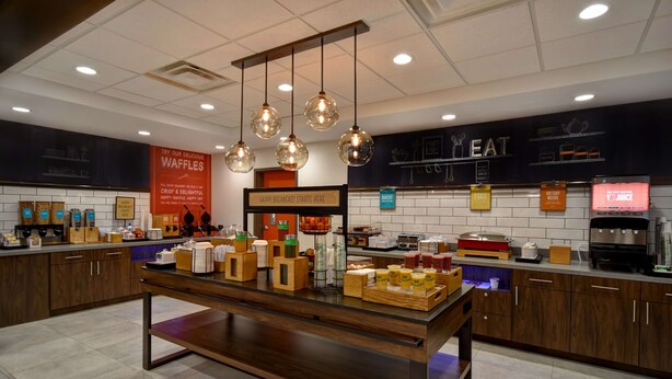 Uma área de café da manhã com mesa de madeira, luminárias pendentes e diversos alimentos expostos no balcão.