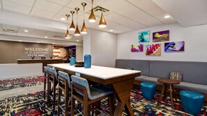 Um saguão de hotel com uma mesa de recepção, área de refeições com uma mesa e cadeiras, um sofá e um tapete estampado.