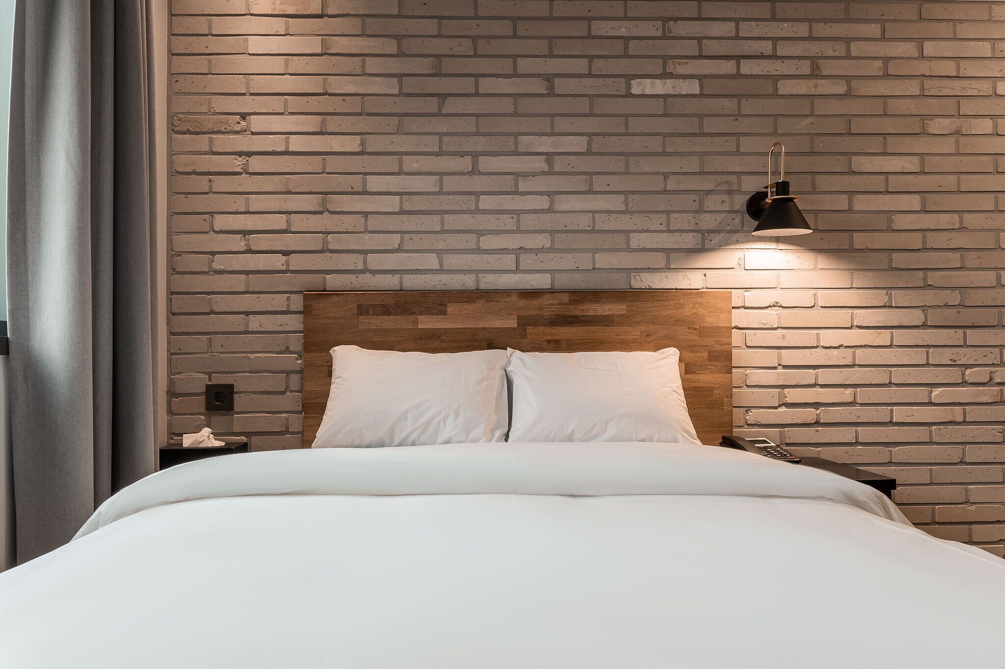 Quarto de hotel com parede em tijolos, cabeceira de madeira, cama branca, mesa de cabeceira com abajur e criado-mudo com telefone.