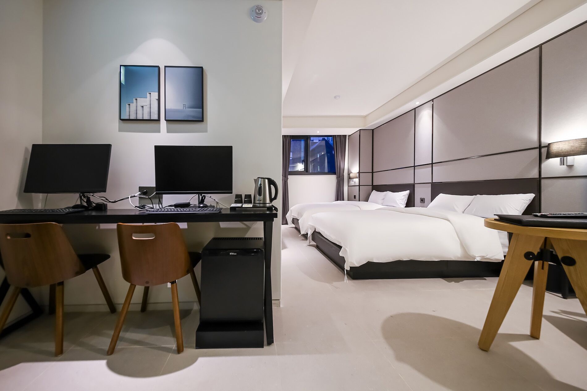 Quarto de hotel moderno com escrivaninha, dois monitores, uma impressora, duas cadeiras, cama com lençóis brancos e uma janela grande com cortinas.
