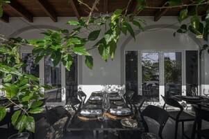Uma área de refeições ao ar livre com uma mesa posta para quatro pessoas, rodeada por cadeiras e uma grande janela com vista para o exterior.