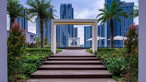 Uma passagem urbana moderna que leva a uma construção emoldurada por edifícios altos, cercada por vegetação exuberante.