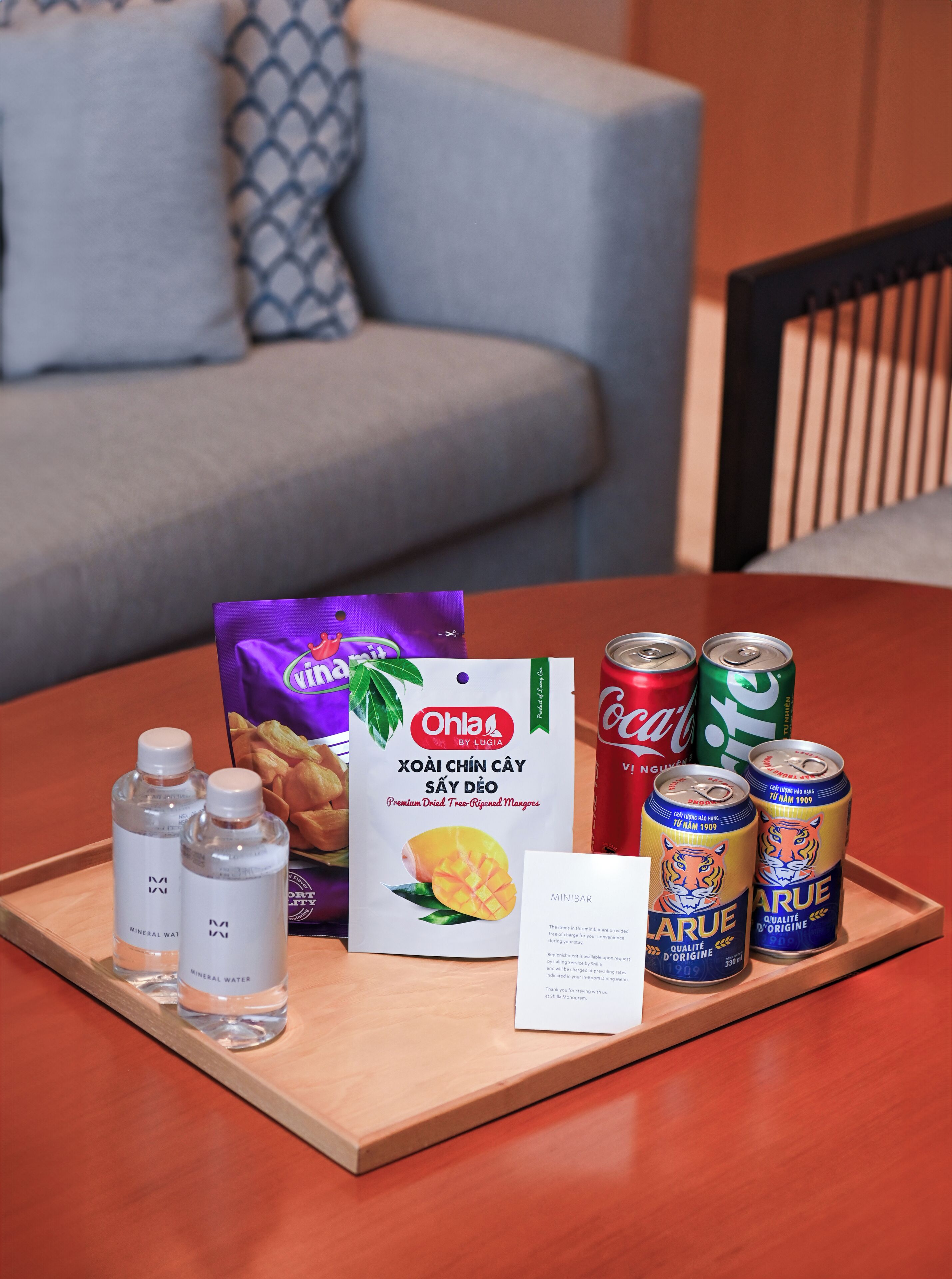 Uma bandeja com garrafas de água mineral, uma caixa de suco de manga Onla, e latas de Coca-Cola e cerveja Tiger em uma mesa de madeira.
