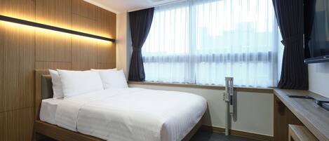Quarto de hotel moderno com uma cama grande, painéis de madeira, televisão de tela plana e uma janela com cortinas translúcidas.