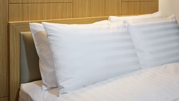 Cama bem arrumada com lençóis e almofadas brancos, cabeceira de madeira e uma faixa de luz horizontal acima.