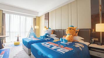 Quarto de hotel com duas camas, cada uma com colcha azul estampada com um personagem de desenho animado. Há uma mesa pequena com abajur, uma janela com cortinas e uma mesa de cabeceira com abajur.