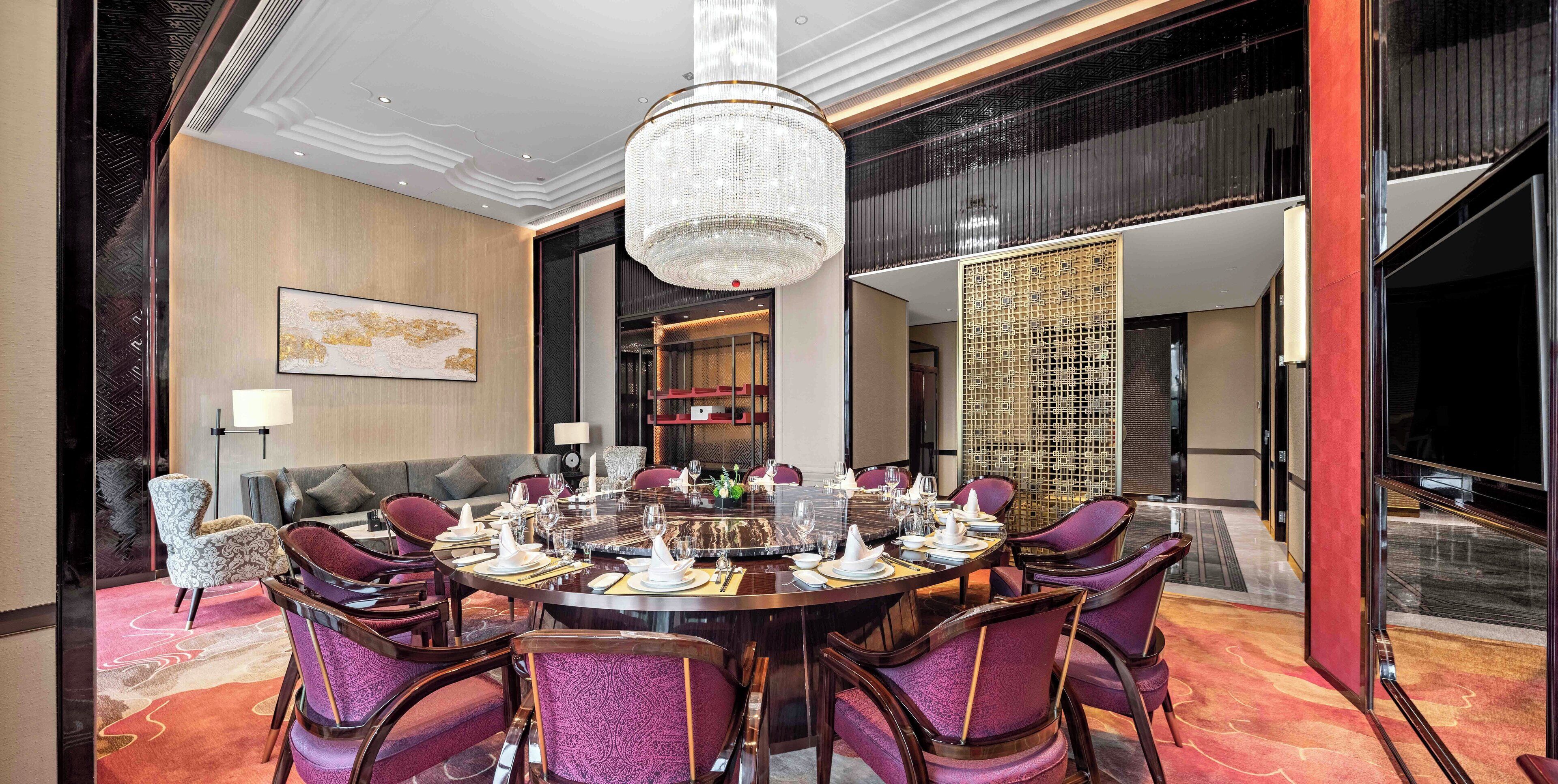 Uma sala de jantar com uma mesa redonda posta para seis pessoas, rodeada por cadeiras estofadas em tons de roxo.